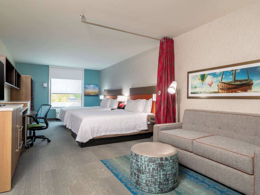 Quarto de hotel com cama, escrivaninha com computador, sofá e um pequeno banquinho redondo.