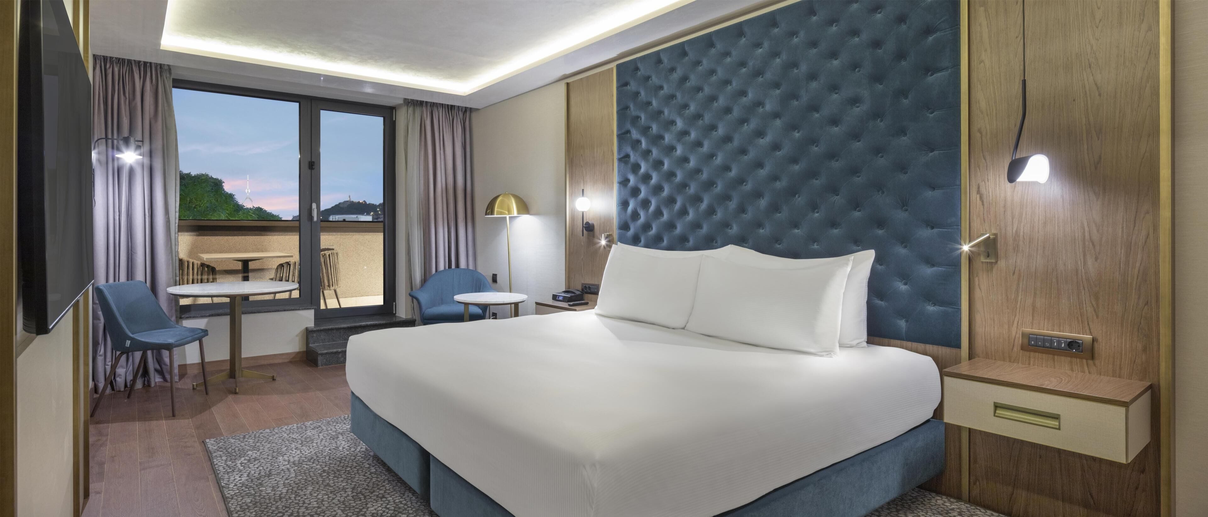 Quarto de hotel com uma cama grande, uma escrivaninha, duas cadeiras, uma mesinha e vista para o exterior.