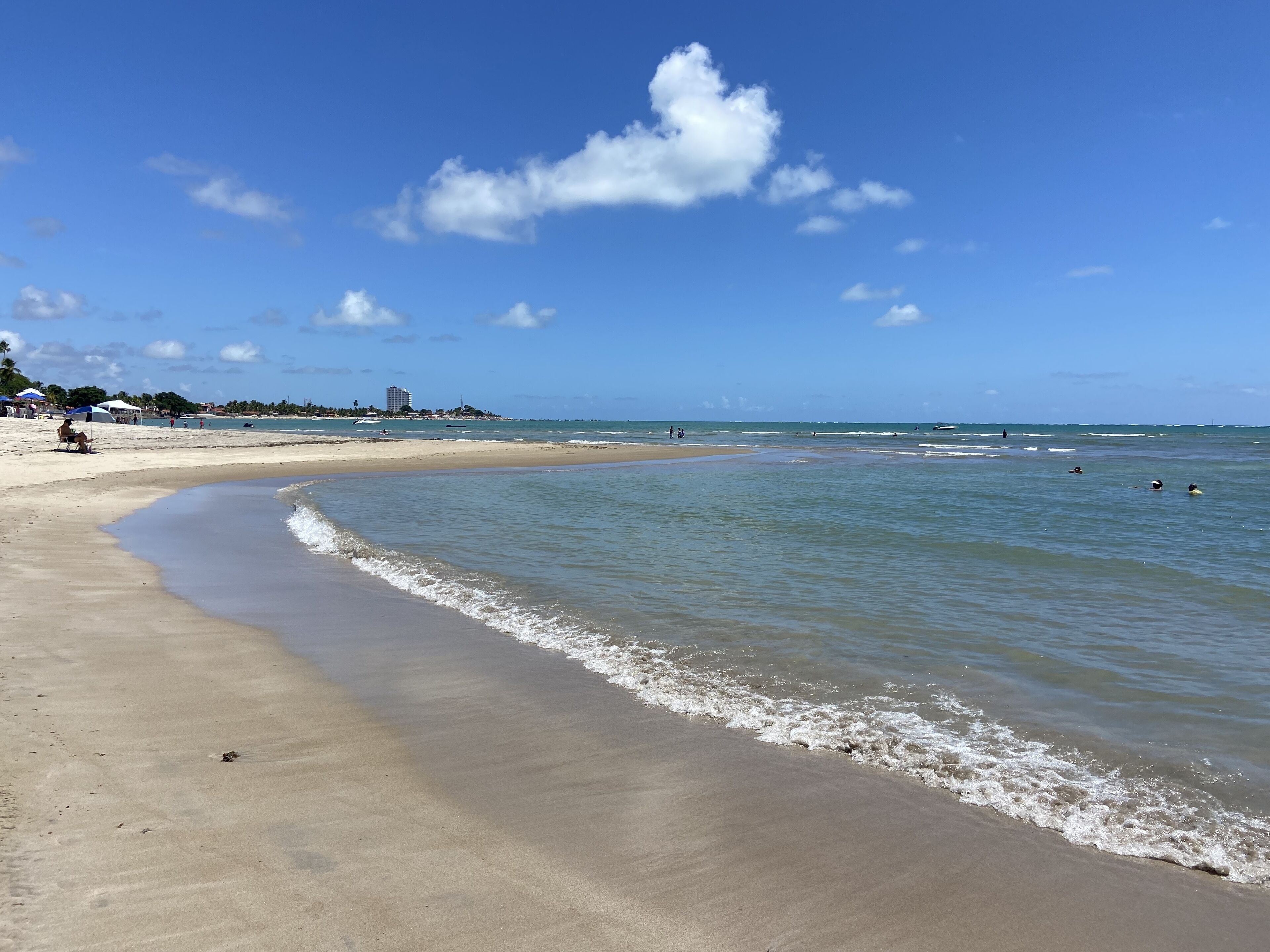 Uma praia com águas azul-claras, margem arenosa e algumas pessoas ao longe.
