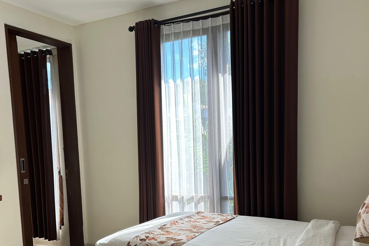 Quarto de hotel com duas camas, uma janela grande com cortinas, uma porta que dá para outro cômodo e um espelho na parede.