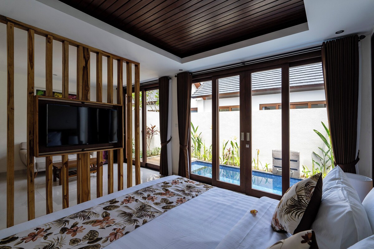 Quarto com uma cama grande, uma televisão fixada em uma divisória de madeira e vista para uma piscina ao ar livre.
