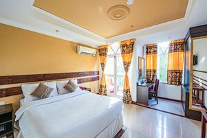 Quarto de hotel com uma cama grande, uma mesa e uma cadeira. Há uma janela com cortinas e vista para vegetação lá fora.