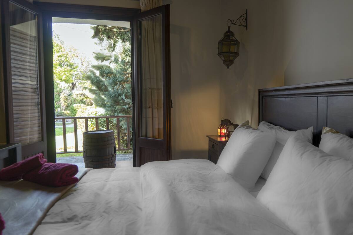 Quarto com cama grande, mesa de cabeceira com lanterna e vista para uma varanda com árvores.