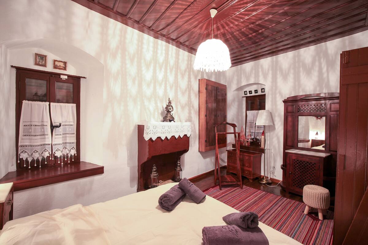 Quarto com cama, cômoda de madeira, espelho, um banquinho pequeno e janela com cortinas.