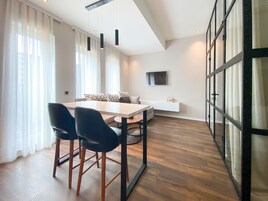 Uma sala de estar moderna com área de jantar, com uma janela grande com cortinas leves, um sofá branco, uma TV na parede e piso de madeira.