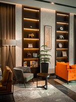 Uma sala de estar moderna com sofá, poltrona e abajur. Prateleiras com objetos decorativos e um vaso de planta também são visíveis.
