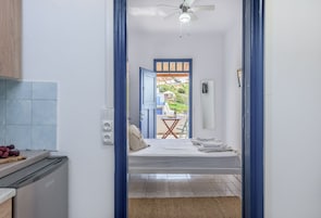 Um corredor estreito leva a um quarto com uma cama, uma mesinha e uma cadeira. Há um ventilador de teto sobre a cama e uma varanda com mesa e cadeiras à vista.