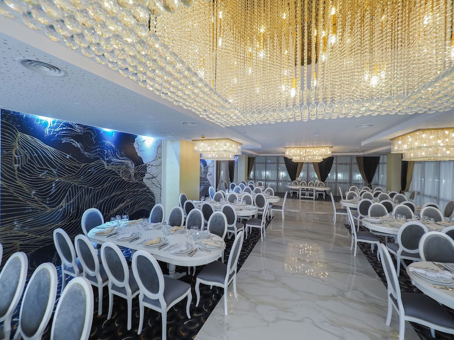 Uma ampla sala de jantar com várias mesas redondas e cadeiras, elegantes lustres e um grande mural na parede.