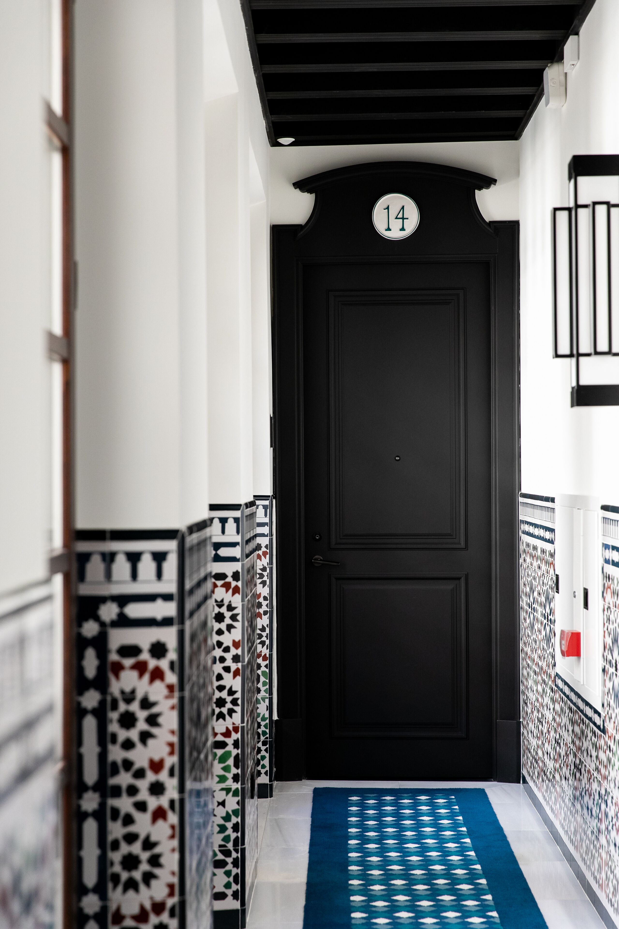Um corredor com uma porta preta marcada com o número 14, grades com padrão e um tapete azul estampado.