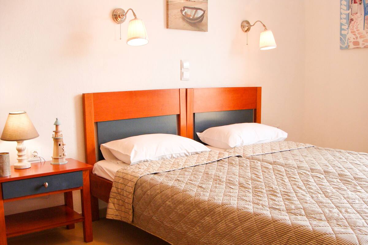 Uma cama bem arrumada com cobertura acolchoada, uma mesa de cabeceira de madeira com abajur e duas luminárias fixadas na parede.