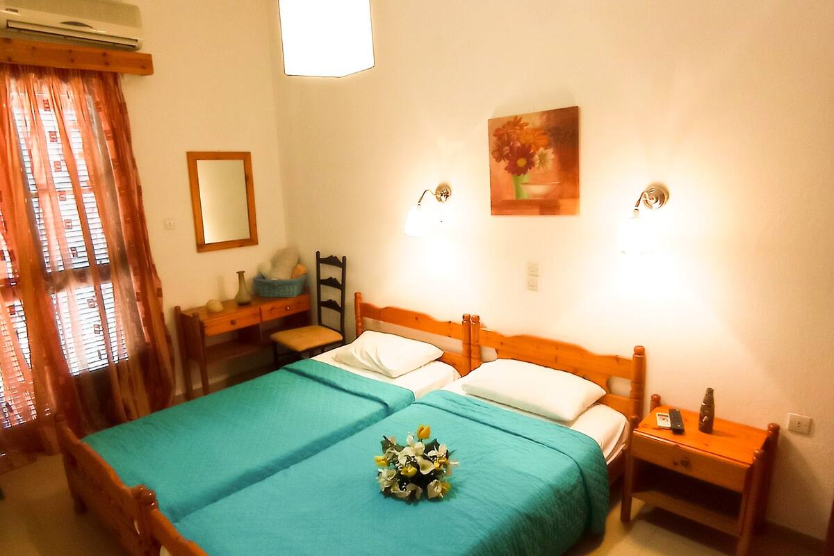 Uma cama de casal com colcha verde-água, mesa de cabeceira com luminária, criado-mudo e um quadro na parede.