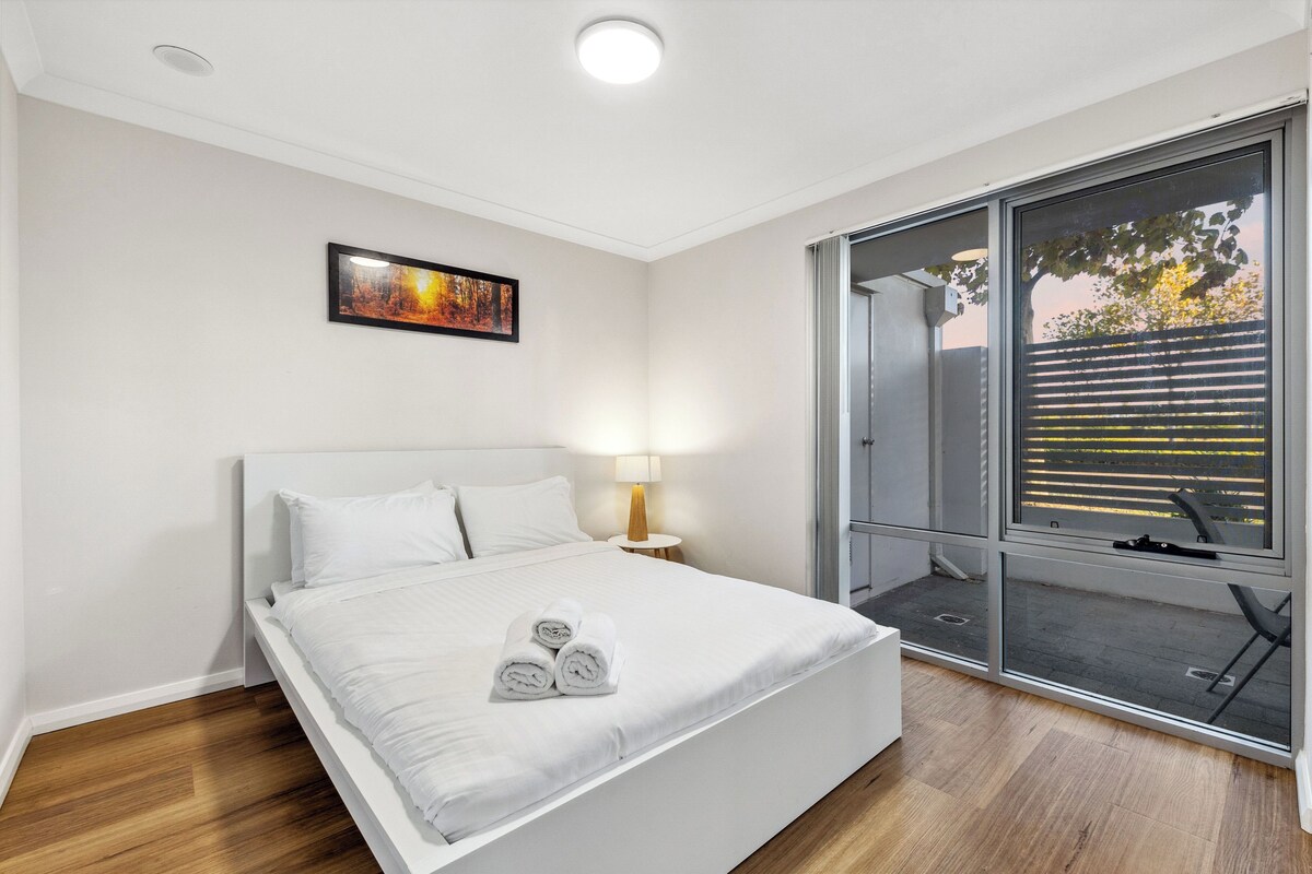 Quarto moderno com cama grande, mesa de cabeceira, abajur e porta de vidro deslizante para área externa.