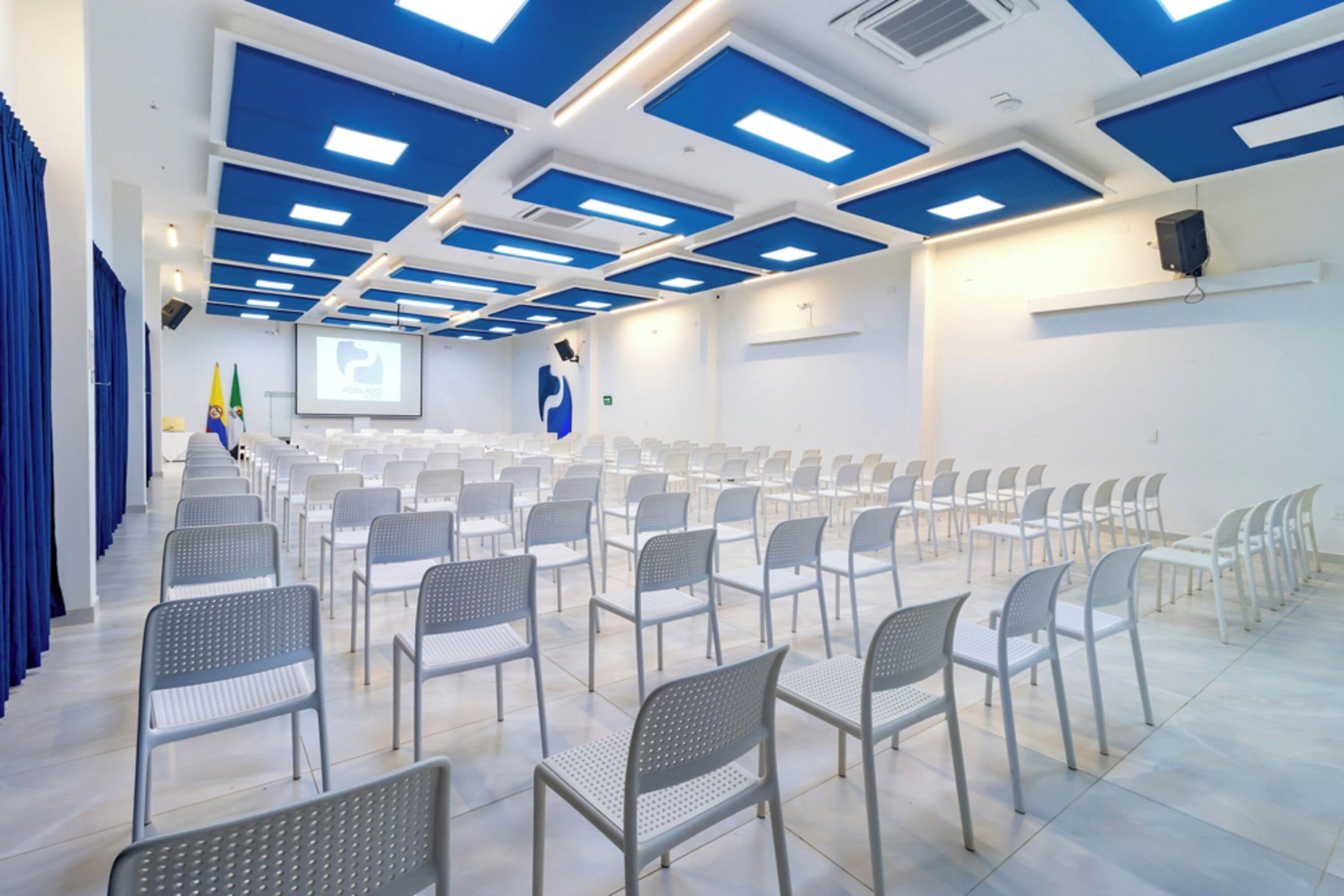 Sala de conferências com fileiras de cadeiras, uma tela de projetor e um design de teto azul e branco.