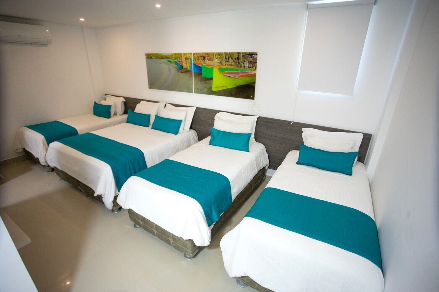 Quarto de hotel com quatro camas individuais, cada uma com roupa de cama branca e verde-água. Há uma janela grande no lado direito e um quadro de barcos na parede acima das camas.