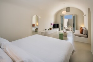 Quarto de hotel moderno com uma cama grande, área de estar com sofá e poltronas, uma mesinha e uma televisão.