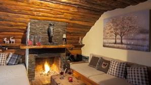 Interior acolhedor de uma cabana de toras, com uma lareira de pedra, um sofá com almofadas xadrez e um quadro de uma árvore na parede.