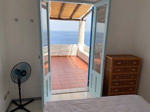 Quarto com uma cama, penteadeira de madeira, ventoinha e porta aberta para uma varanda com vista para o mar.