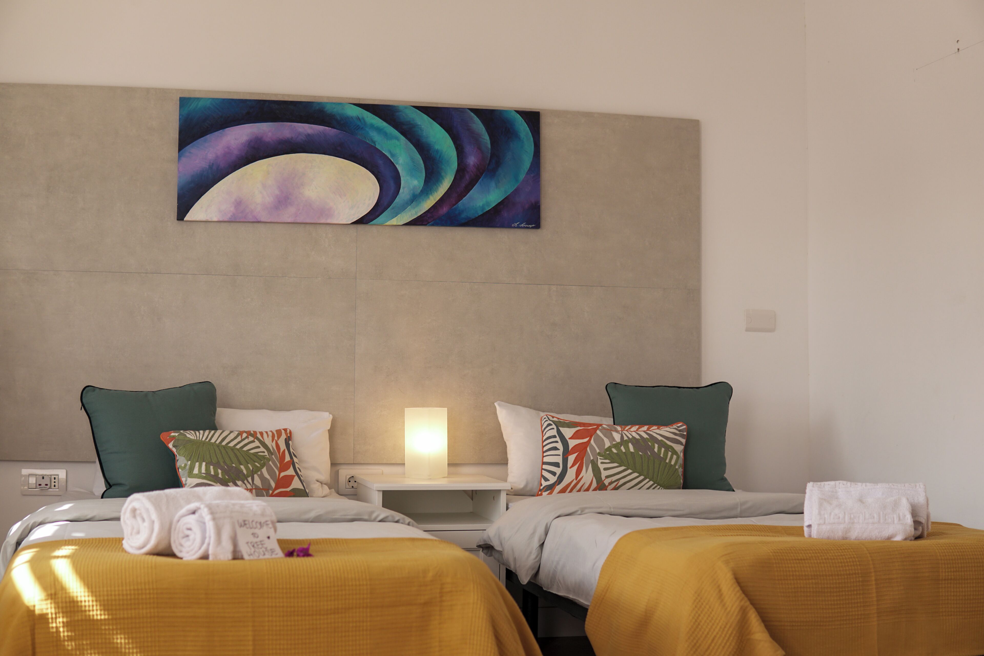 Quarto de hotel com duas camas, uma mesinha de cabeceira, um quadro na parede e um abajur.