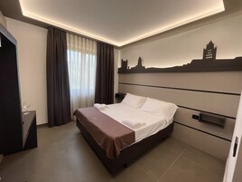 Quarto de hotel moderno com uma cama grande, mesa de cabeceira, espelho e arte na parede retratando uma paisagem urbana.
