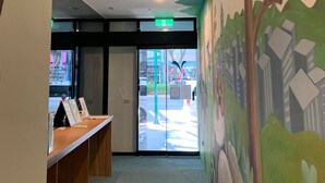 Corredor interno com um mural de uma árvore e uma coruja, uma mesa de recepção e uma porta de vidro que dá para o exterior.