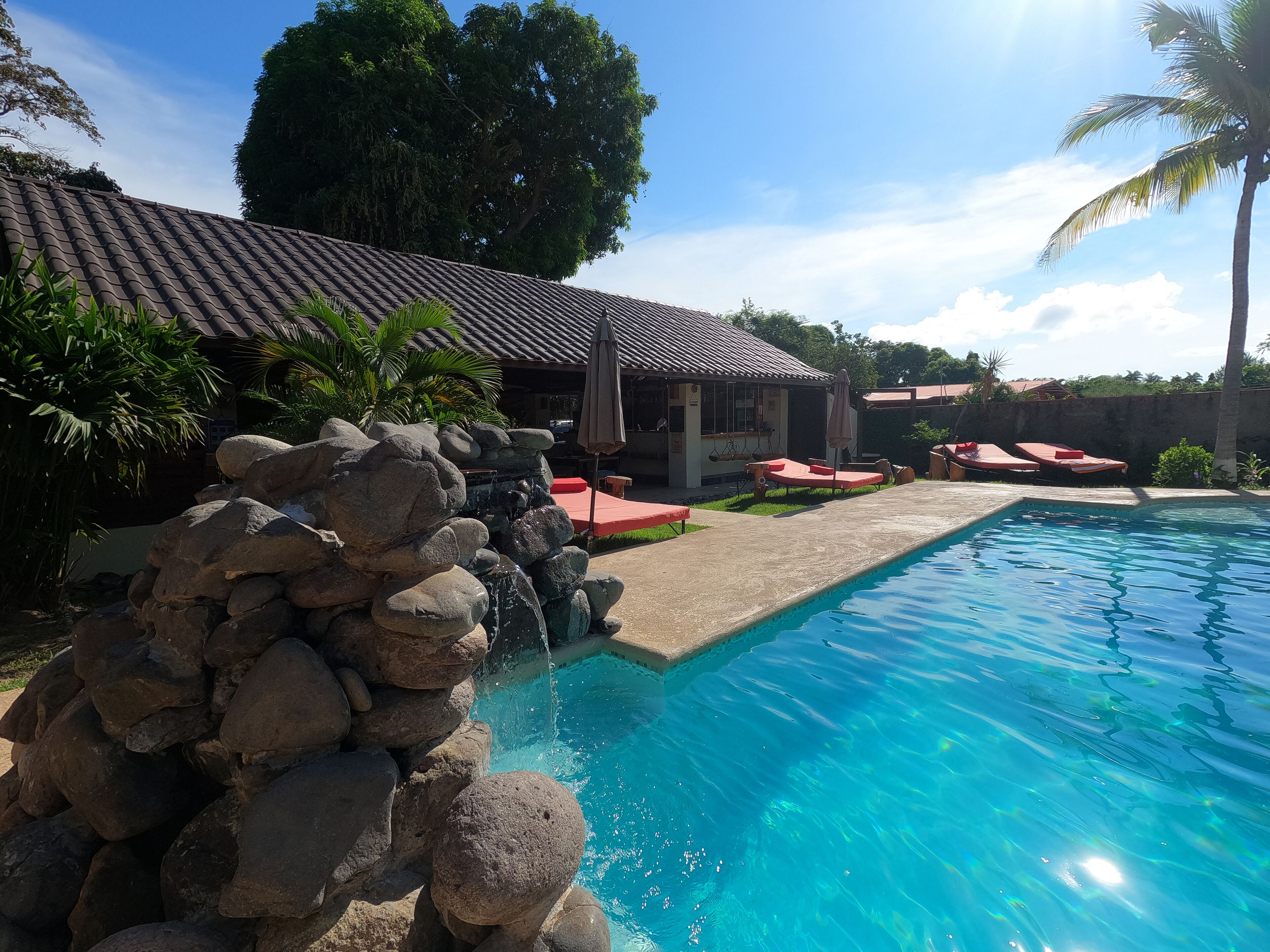 Uma piscina com uma parede de pedra em primeiro plano, uma área de estar coberta com almofadas vermelhas e um edifício com telhado telhado ao fundo.