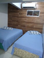 Quarto com duas camas, cada uma com colcha azul e almofadas listradas, parede de madeira, ar condicionado e janela.