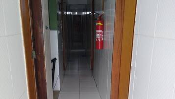 Um corredor com paredes revestidas de azulejos brancos, um extintor preso na parede e uma porta à direita.