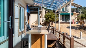 Uma área de estar coberta ao ar livre, com uma mesa e cadeiras de madeira, guarda-corpo de metal e vista para um bairro residencial com casas e uma árvore.