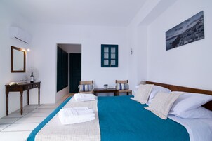 Quarto de hotel com uma cama grande, mesa de cabeceira com garrafa e copos, uma cadeira, uma mesinha e um quadro na parede.