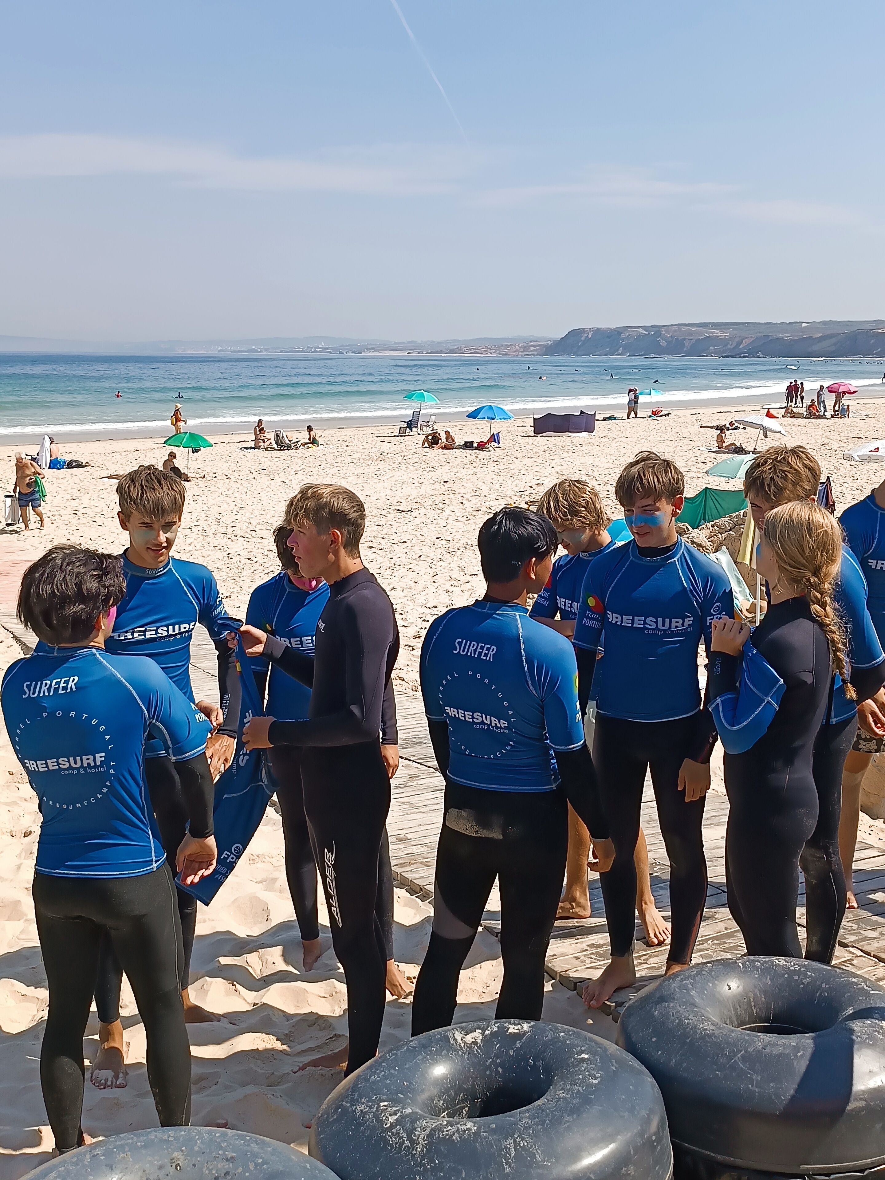 Um grupo de pessoas em fatos de mergulho azuis com os logotipos 'SURFER' e 'FREESURF', em pé numa praia com o oceano ao fundo.