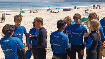 Um grupo de pessoas em fatos de mergulho azuis com os logotipos 'SURFER' e 'FREESURF', em pé numa praia com o oceano ao fundo.