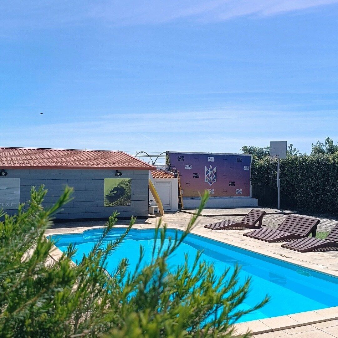 Piscina com espreguiçadeiras, um pequeno edifício com um mural e um grande outdoor.