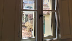 Quarto com uma grande janela, paredes brancas e vista externa para edifícios e uma árvore.