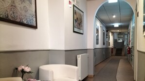 Um corredor com uma poltrona branca, uma mesinha com um vaso de flores, um quadro emoldurado e um radiador.