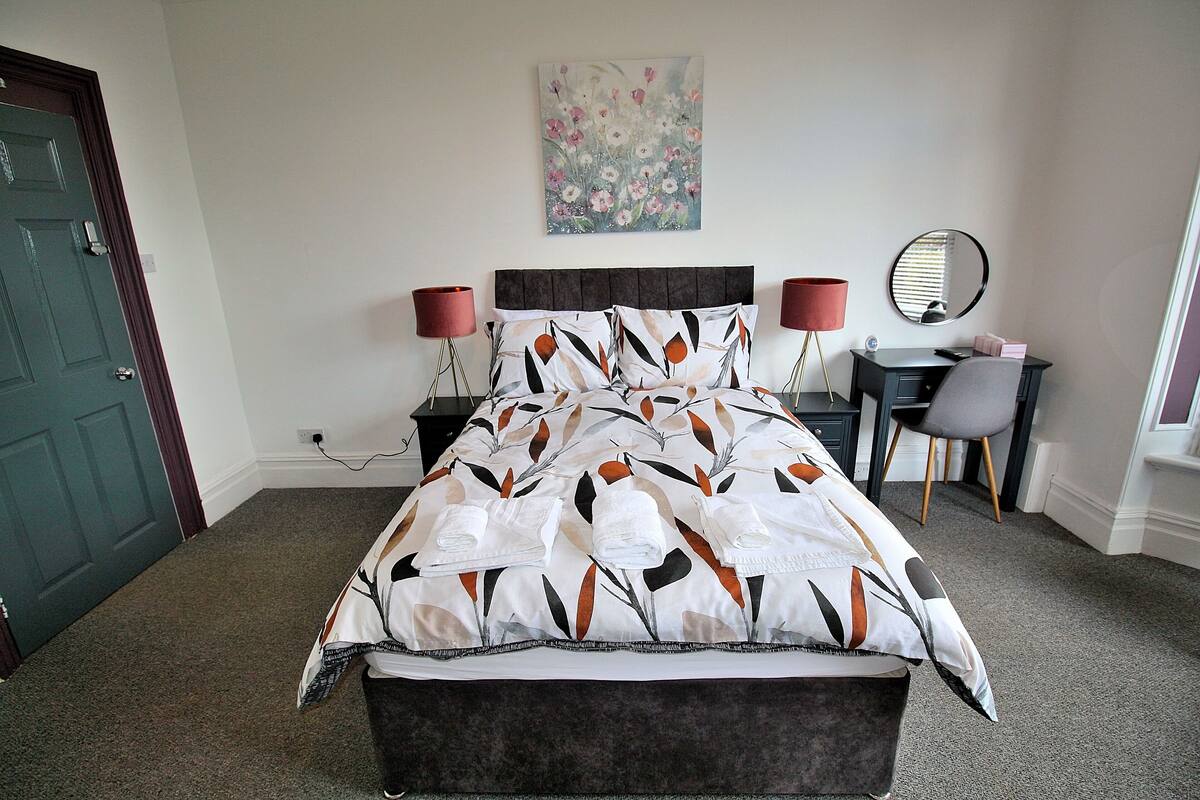 Quarto com cama, escrivaninha, cadeira, espelho e porta.