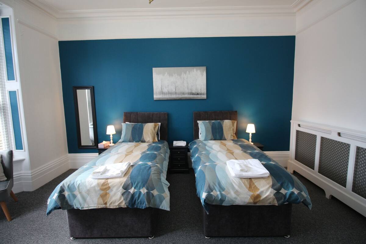 Quarto de hotel com duas camas, um espelho grande, um quadro na parede e uma janela com persianas.