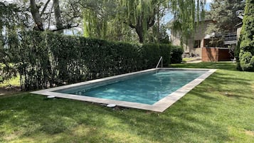 Piscina retangular com escada de metal, rodeada por grama bem cuidada e sebes.