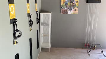 Quarto com teto branco, parede cinzenta e carpete cinza. Há tiras pretas penduradas na parede, uma estante de metal branca e um cartaz na parede.