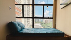 Quarto com cama posicionada sob uma janela ampla, com vista para prédios altos e céu claro.