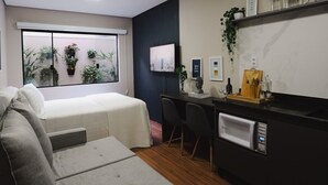 Quarto de hotel compacto com uma cama, escrivaninha, cadeira, um pequeno sofá, janela com plantas, televisão e micro-ondas.