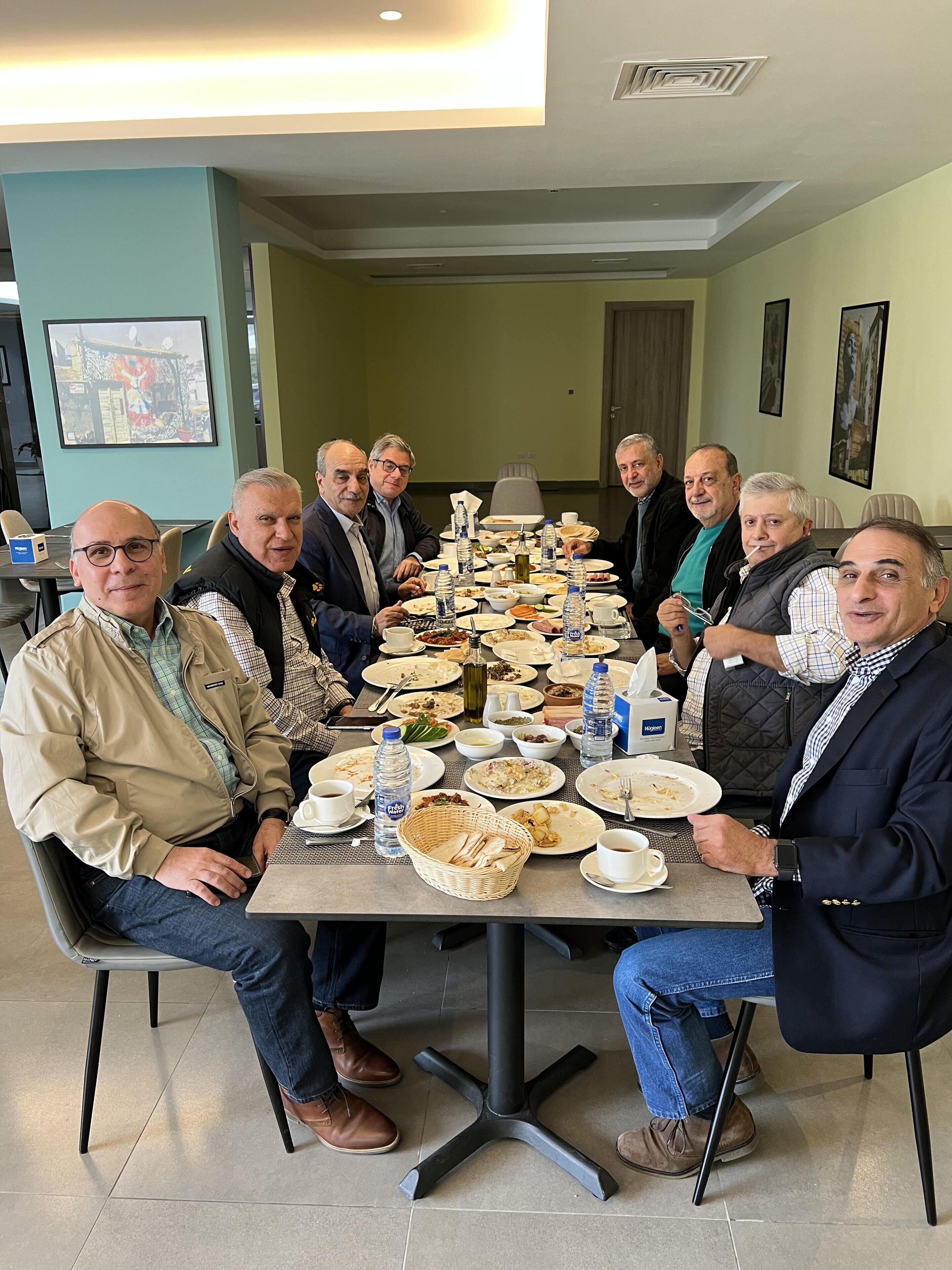 Um grupo de pessoas sentadas à volta de uma mesa, com vários pratos e bebidas, numa sala com chão em mosaico e um quadro na parede.