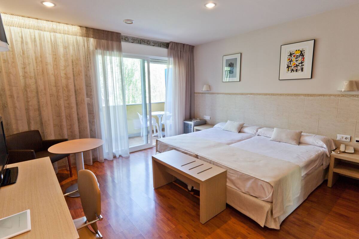Quarto de hotel com uma cama grande, uma escrivaninha, uma cadeira, uma mesinha e varanda com cadeira.