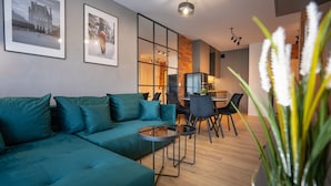Uma sala de estar moderna com um sofá azul-esverdeado, uma área de jantar com mesa e cadeiras, e uma cozinha ao fundo.