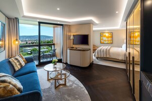 Quarto de hotel moderno com sofá, mesa de centro redonda, cama, televisão e vista para a cidade.