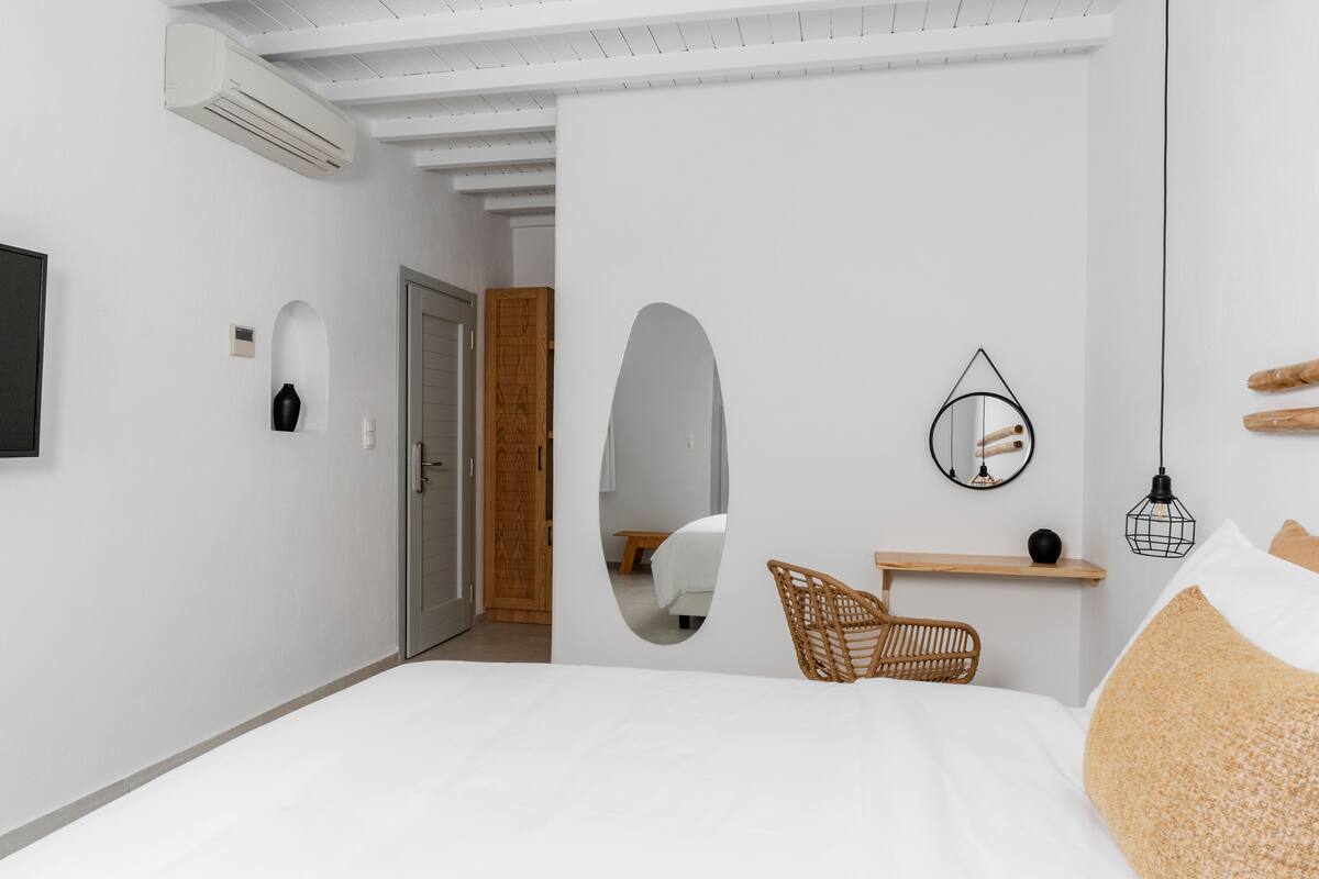 Quarto moderno com uma cama grande, porta de madeira, espelho e um pequeno espaço de estar.