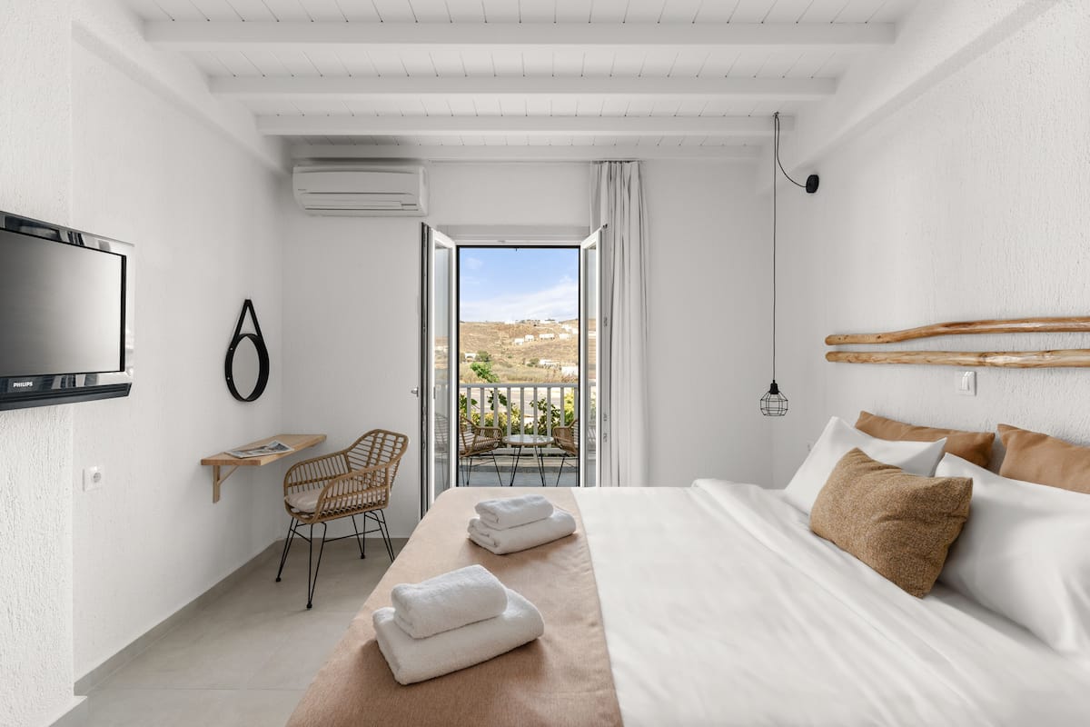 Quarto de hotel moderno com uma cama grande, televisão fixada na parede, uma cadeira e vista para o exterior através de uma porta aberta.