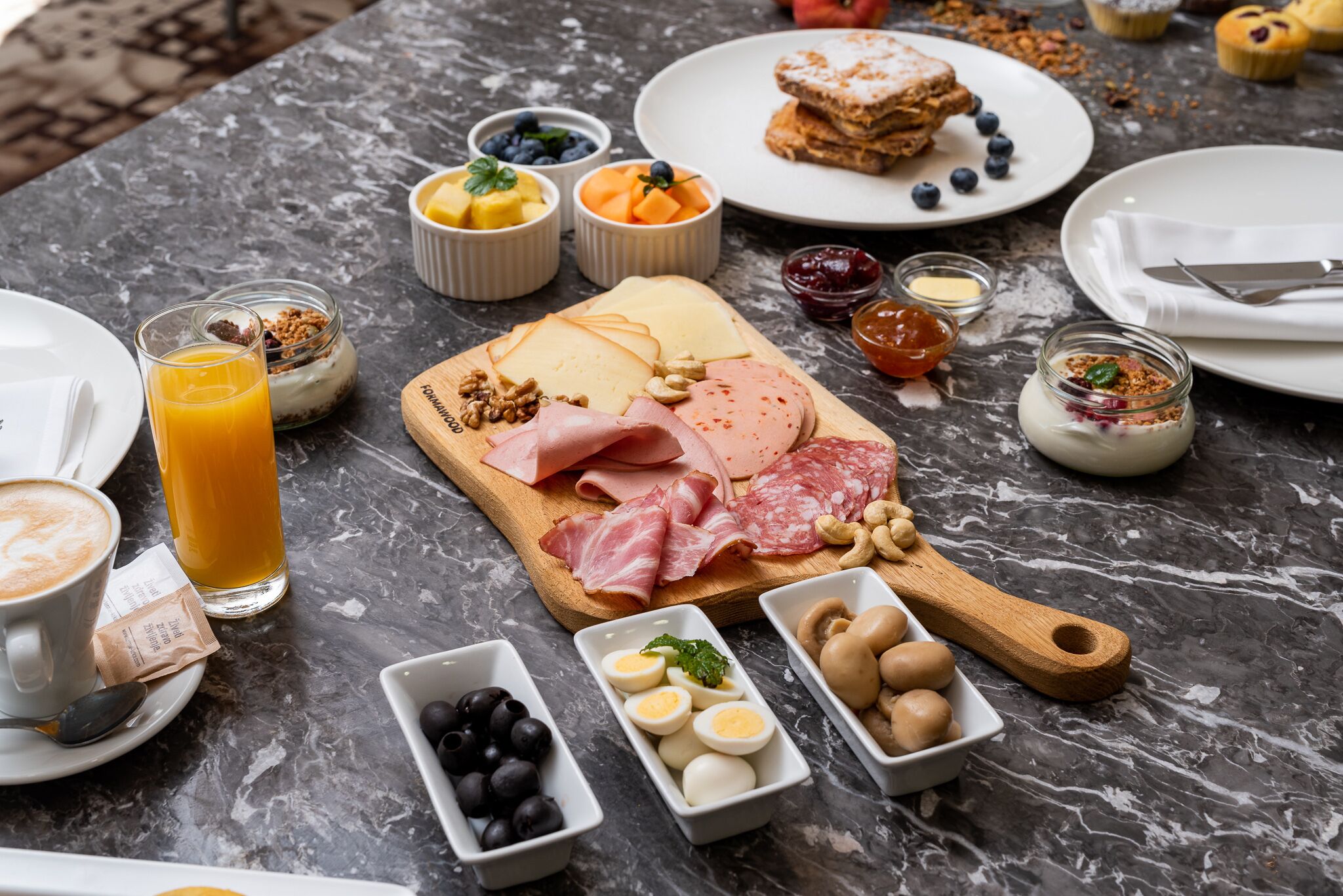 Um buffet de café da manhã com diversos queijos, carnes, frutas e bebidas sobre uma mesa de mármore.