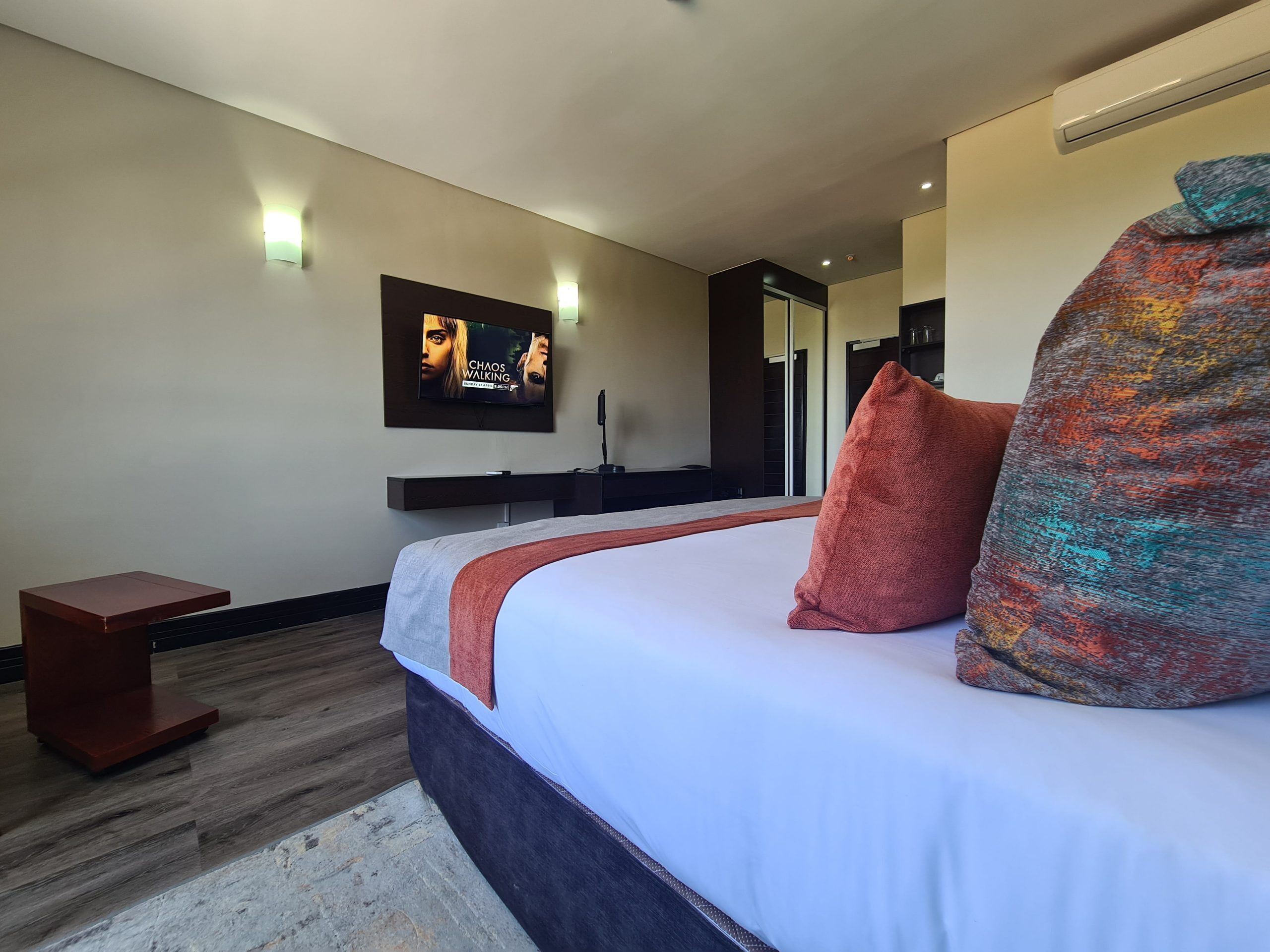 Quarto de hotel moderno com uma cama grande, televisão de tela plana na parede, uma pequena mesa de cabeceira de madeira e uma janela ampla.