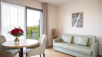 Quarto com uma mesa redonda de madeira, duas cadeiras, um sofá verde claro e uma janela grande com cortinas.