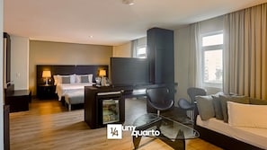 Quarto de hotel com cama, televisão, escrivaninha com computador, mesa de vidro e sofá.